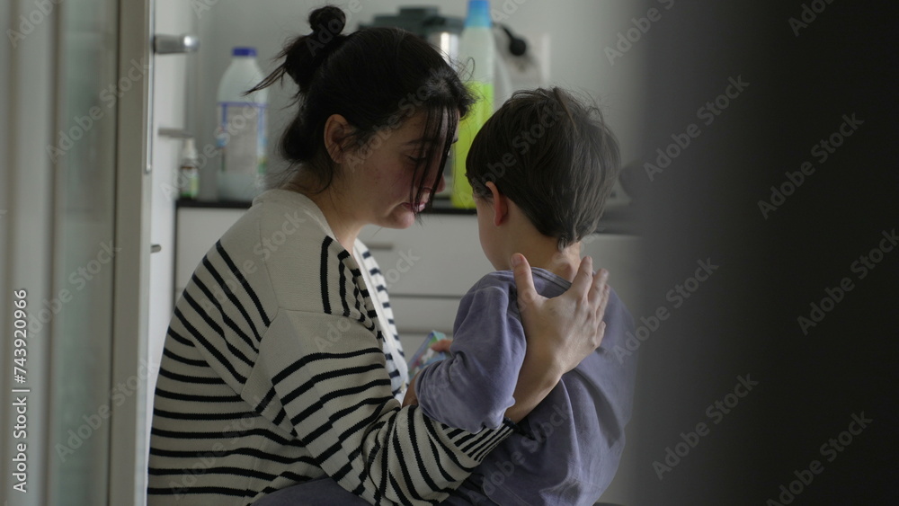 Caring mother consoling crying little son in candid kitchen scene. Mom embracing hurt child ...