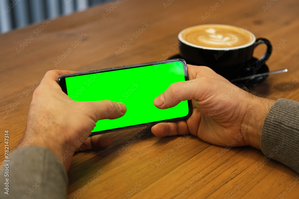 Young man sitting at cafe holding smartphone green mock-up screen in hand. Male person using ...