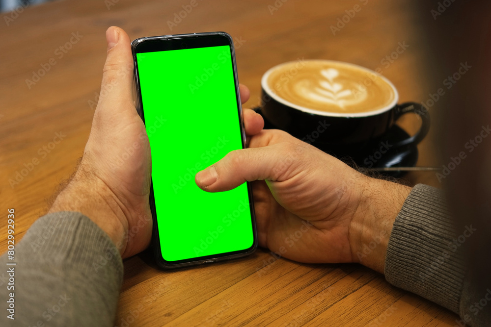 Young man sitting at cafe holding smartphone green mock-up screen in hand. Male person using ...