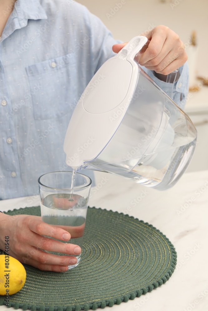 Woman pouring water from filter jug into glass at white marble table in ...