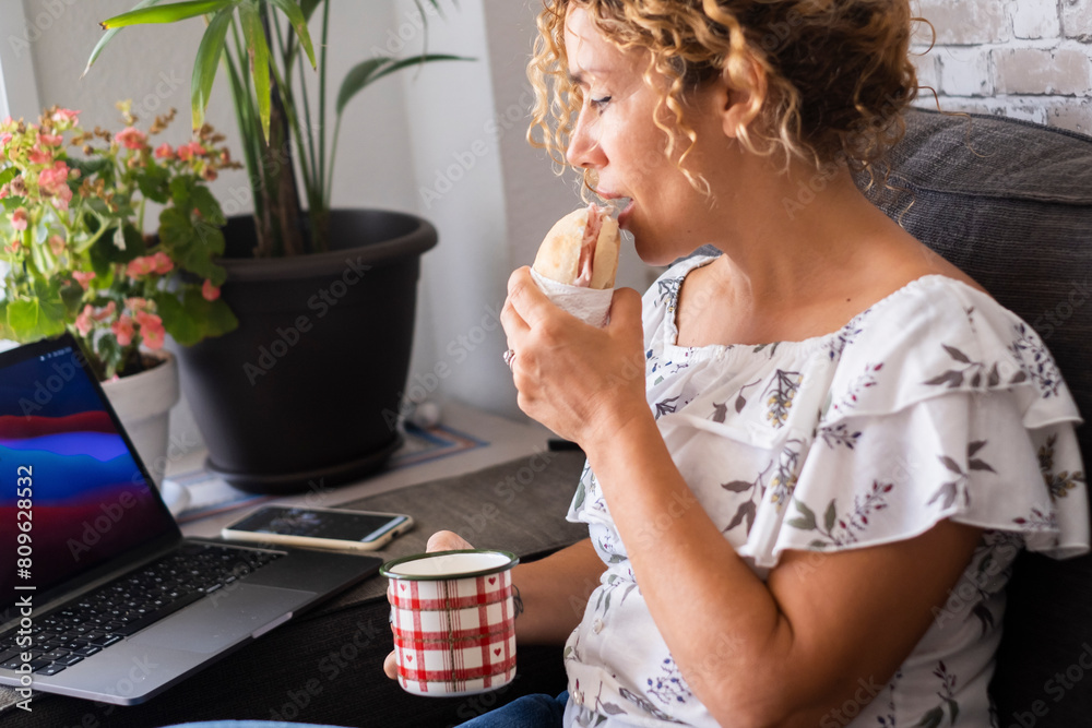 One unhealthy woman eating sandwich and working on laptop at home ...