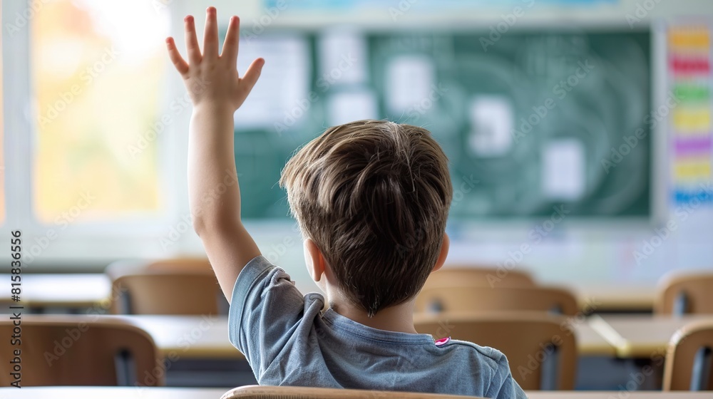 A smart schoolboy sits at a desk in a classroom with his hand raised ...