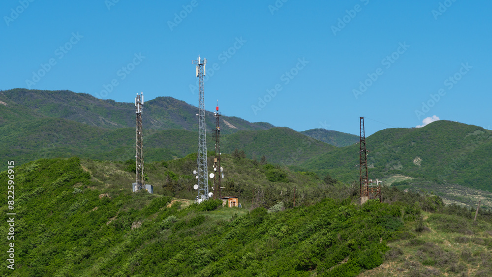 Mobile network antennas are positioned atop a green hill with mountains ...