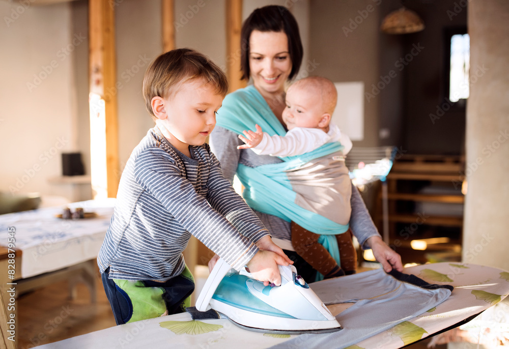 Mother carrying small baby in wrap, older son helping with ironing ...