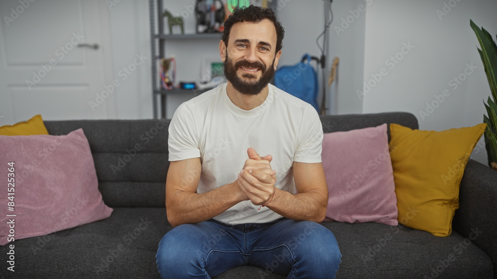 Bearded man smiling casually sits on a grey couch indoors, exuding a ...
