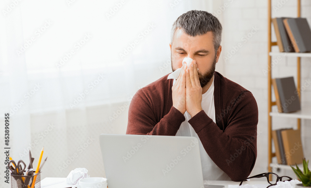 Sick man sitting in front of a laptop is blowing his nose into a tissue ...