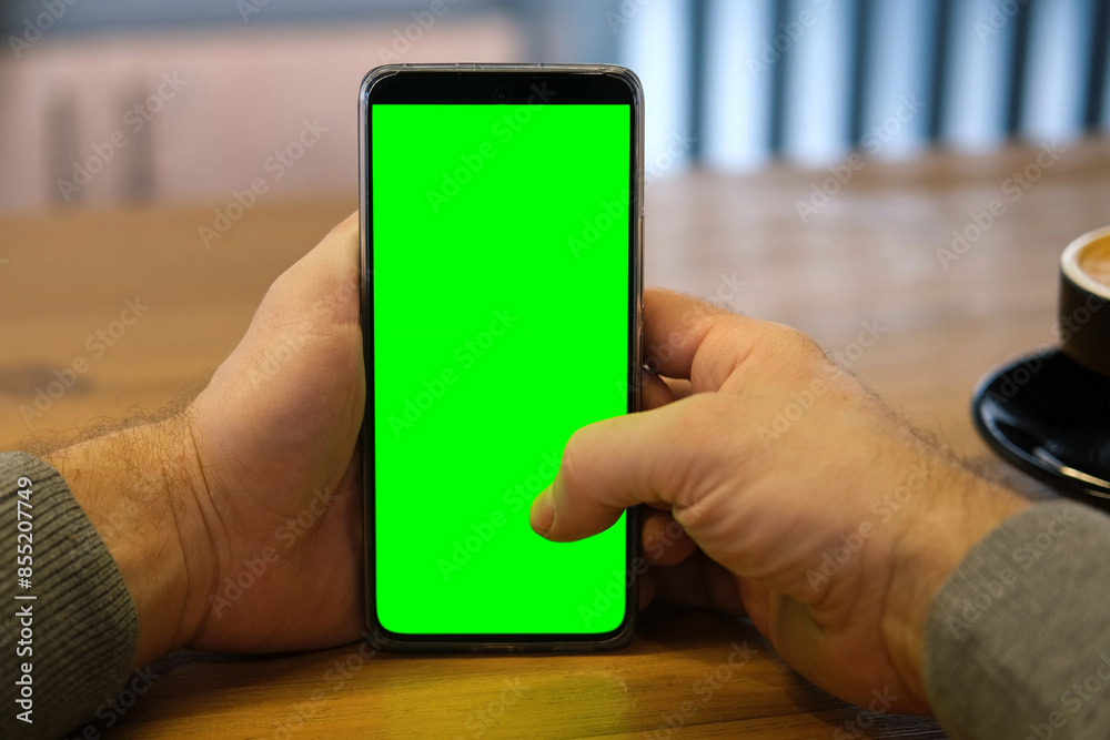 Young man sitting at cafe holding smartphone green mock-up screen in hand. Male person using ...