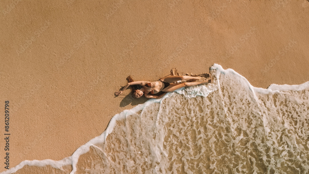 Aerial view of young sexy woman in bikini lying on golden sand beach basking in the sun while ...