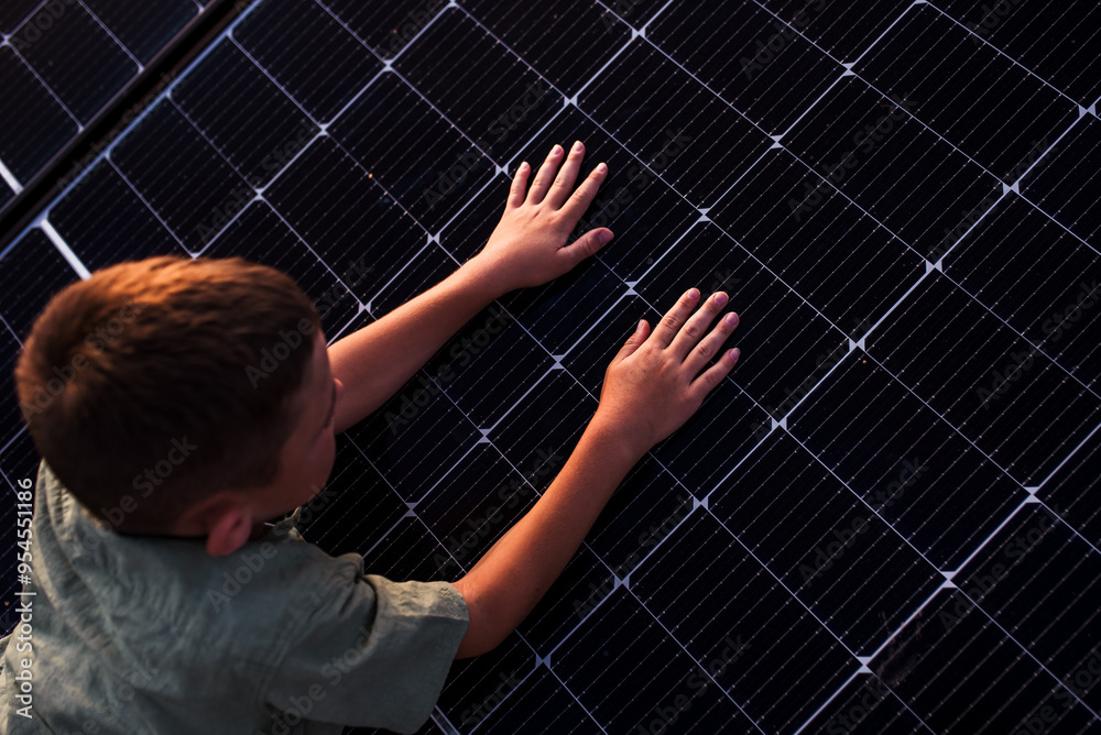Boy lying on belly on roof with solar panels, touching panel with hands ...