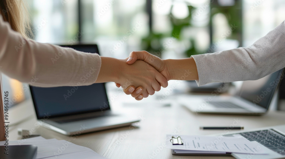 Two businesswomen shake hands firmly over a desk in a modern office ...