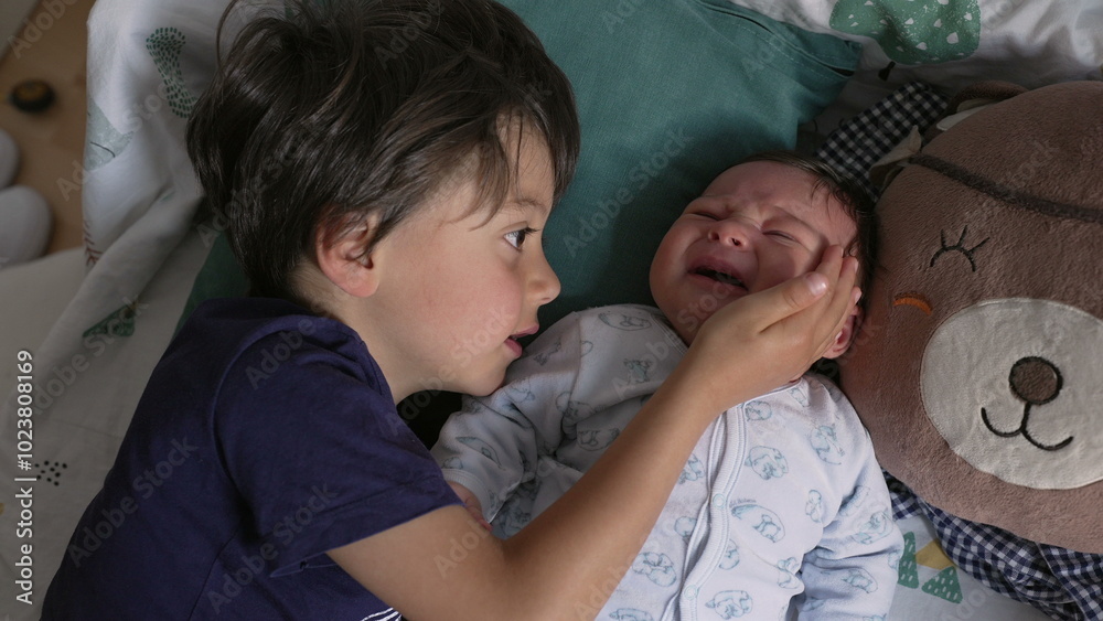 Young boy gently comforting his crying newborn sibling, both lying on a ...