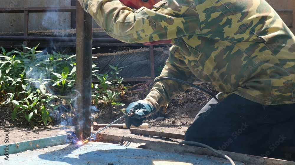 welder in green jacket and red mask welds metal beam to iron base ...