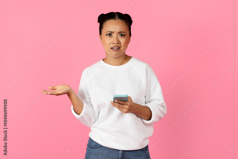Asian woman with a puzzled expression holds her smartphone and gestures ...