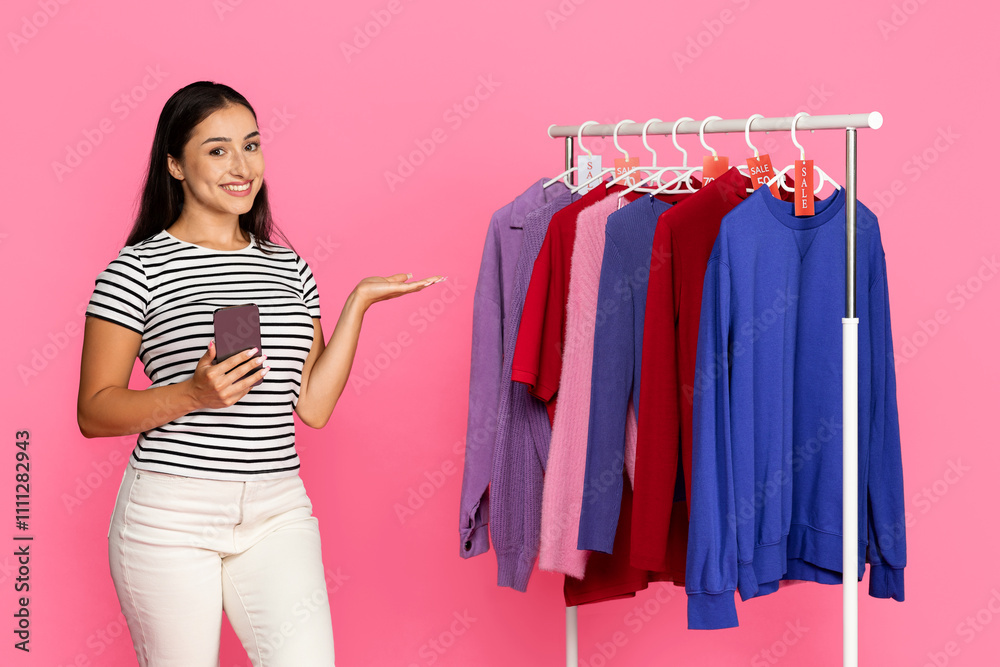 A young woman stands excitedly beside a rack of vibrant sweaters. She ...