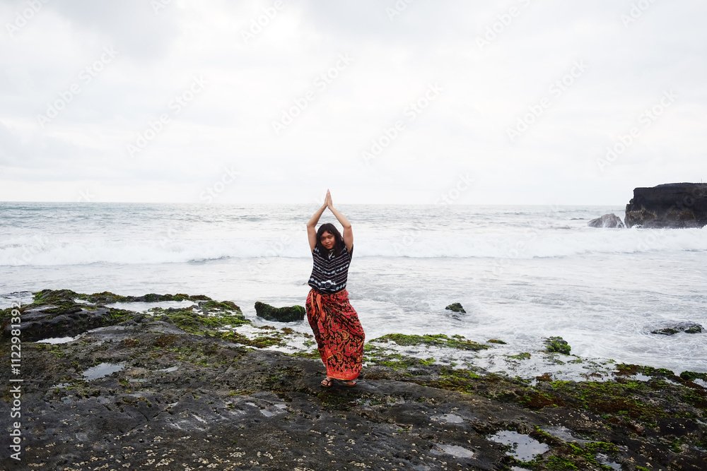 Asian woman wearing native Sarong of Bali doing yoga poses on the coast ...