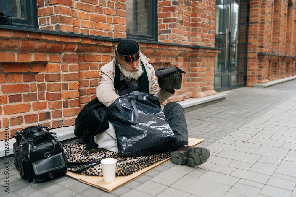 Elderly homeless man in hat sitting outside in cold, holding out cup ...