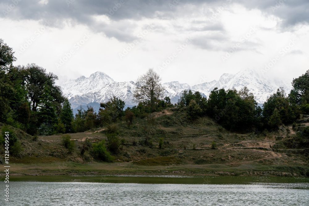 Snow covered mountains rise behind a lush green forest and a calm lake ...