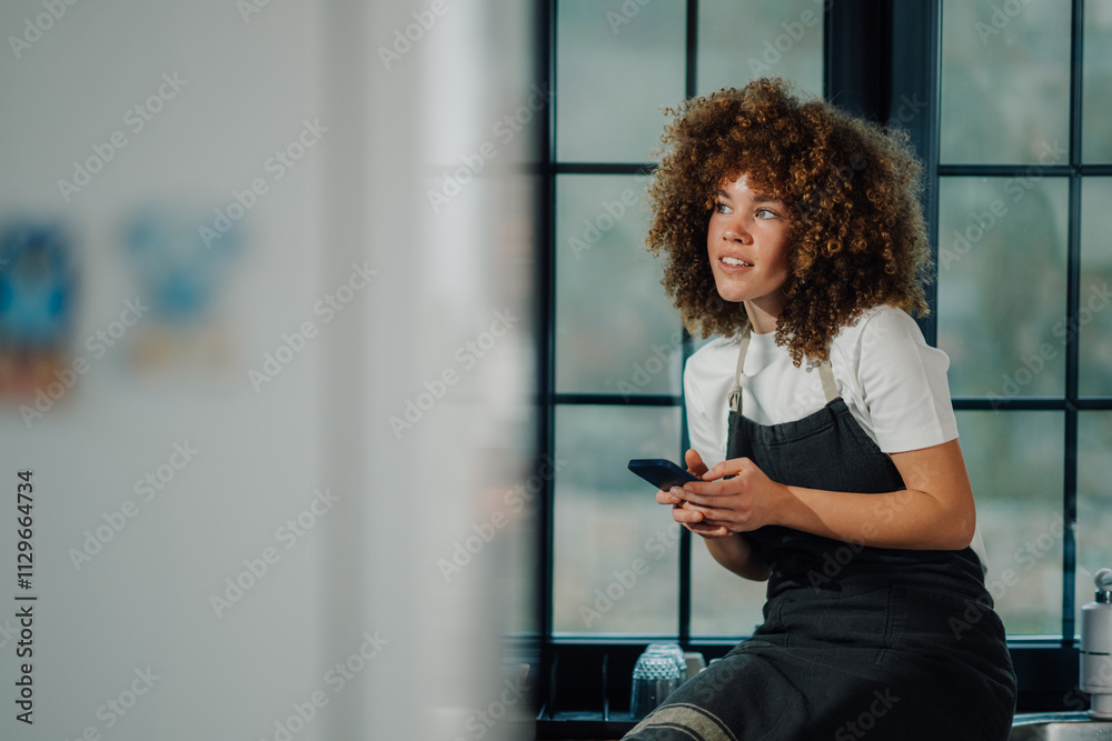Smiling waitress holding smartphone and looking away in restaurant ...