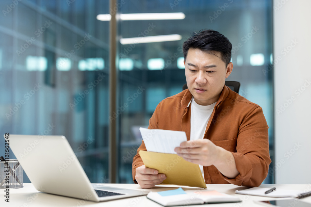 Asian man dressed casually examining documents at a modern office desk ...
