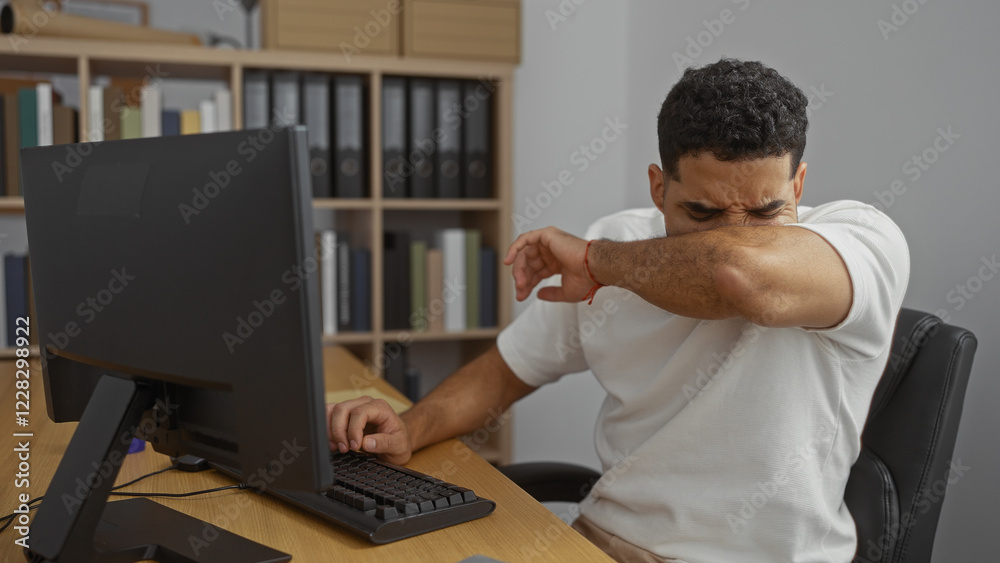 Young man sneezing into his elbow while working at a computer in an ...