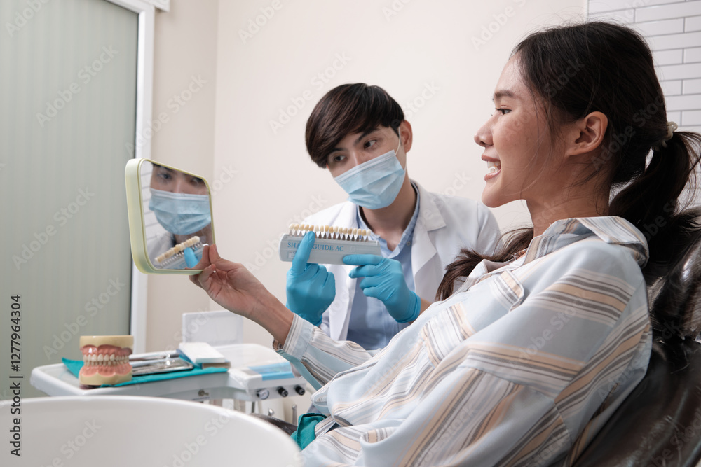 Asian male dentist explains color shades of tooth implant whitening to young female patient ...