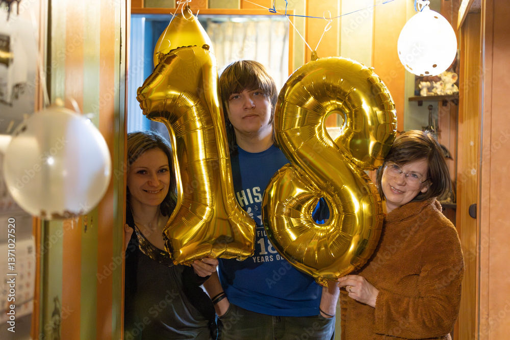 Three people pose indoors with large gold number balloons shaped as 18 ...