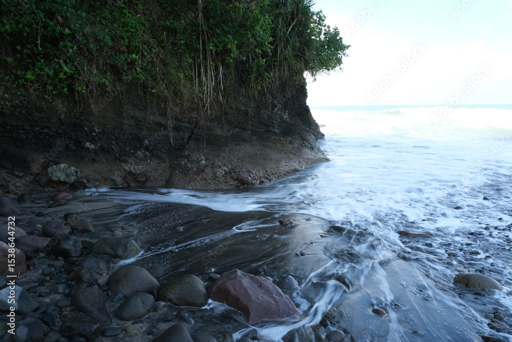 Ocean waves flow over dark rocks and black sand at the base of a lush ...