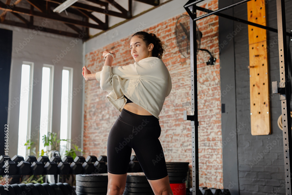Female gym-goer stretching arms in brick-walled gym by windows with ...