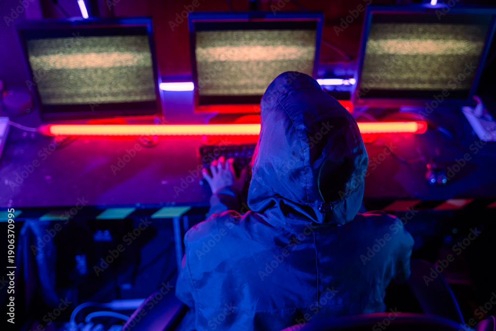 Photo Stock Hooded figure operating at keyboard in dark room facing ...
