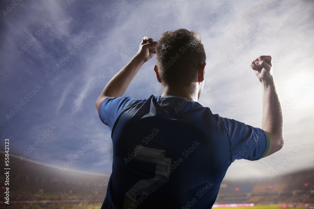 Soccer player with arms raised cheering, stadium with sky and clouds ...