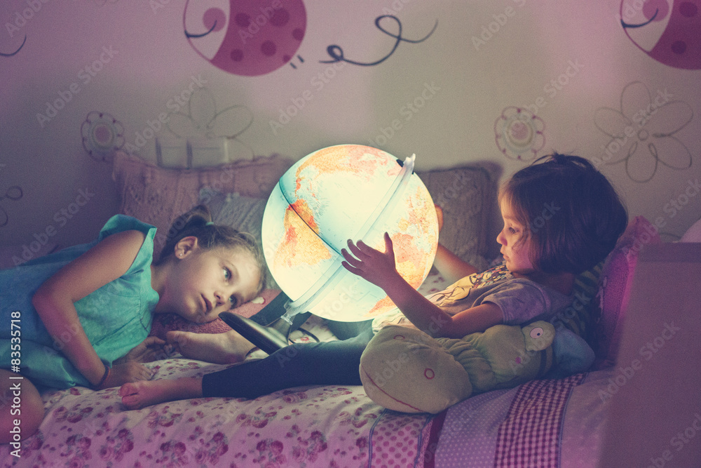 Girls looking at illuminated lamp in shape of globe while lying on bed ...