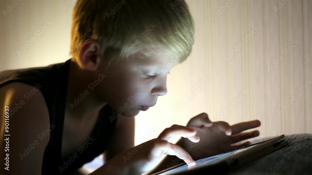 Kid playing computer games laying in his bed in evening using touchpad ...
