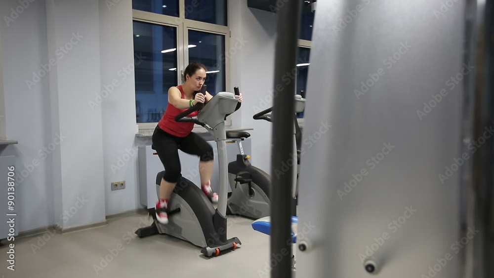 Fat woman at the gym on an exercise bike. Slider camera movement right ...
