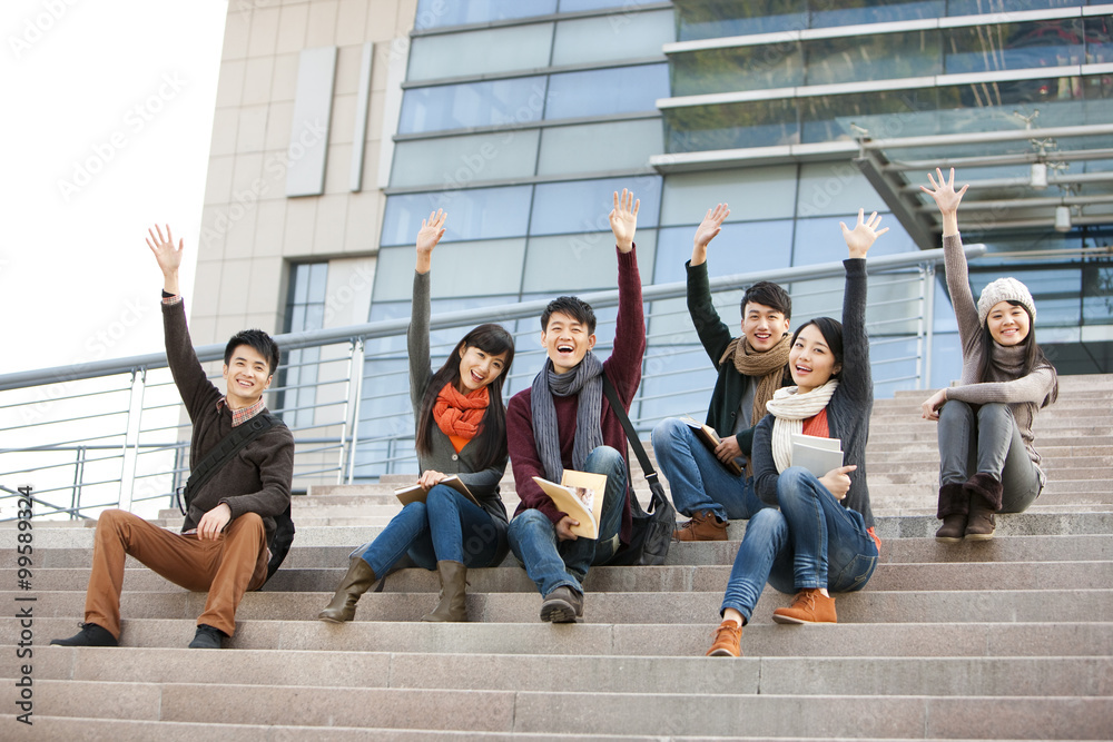Young college students waving and sitting on steps of university ...