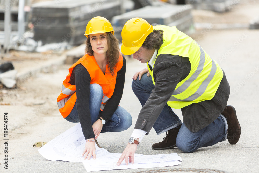 Male and female crouching architects with safety jackets and hardhats ...