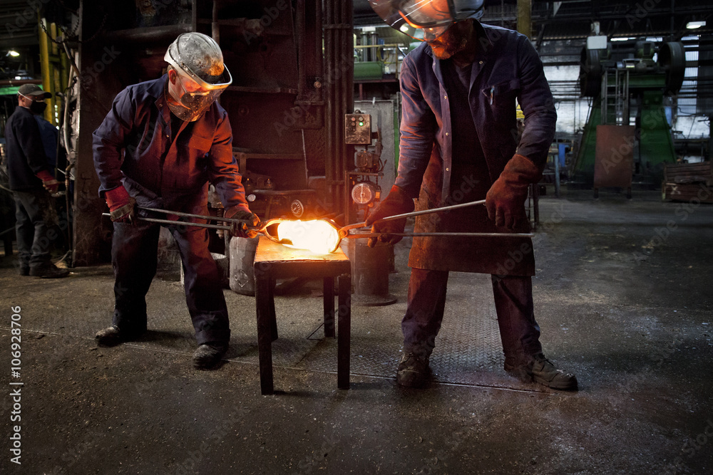 Forge worker hands over pre formed steel billet to colleague for ...