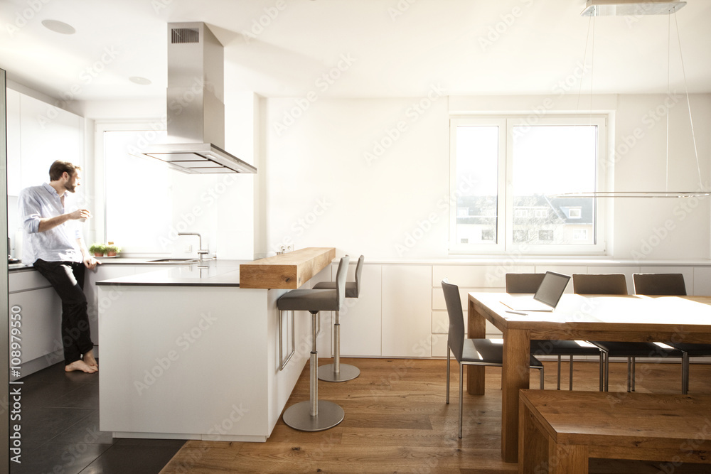Man with coffee cup standing in his open plan kitchen, looking through ...