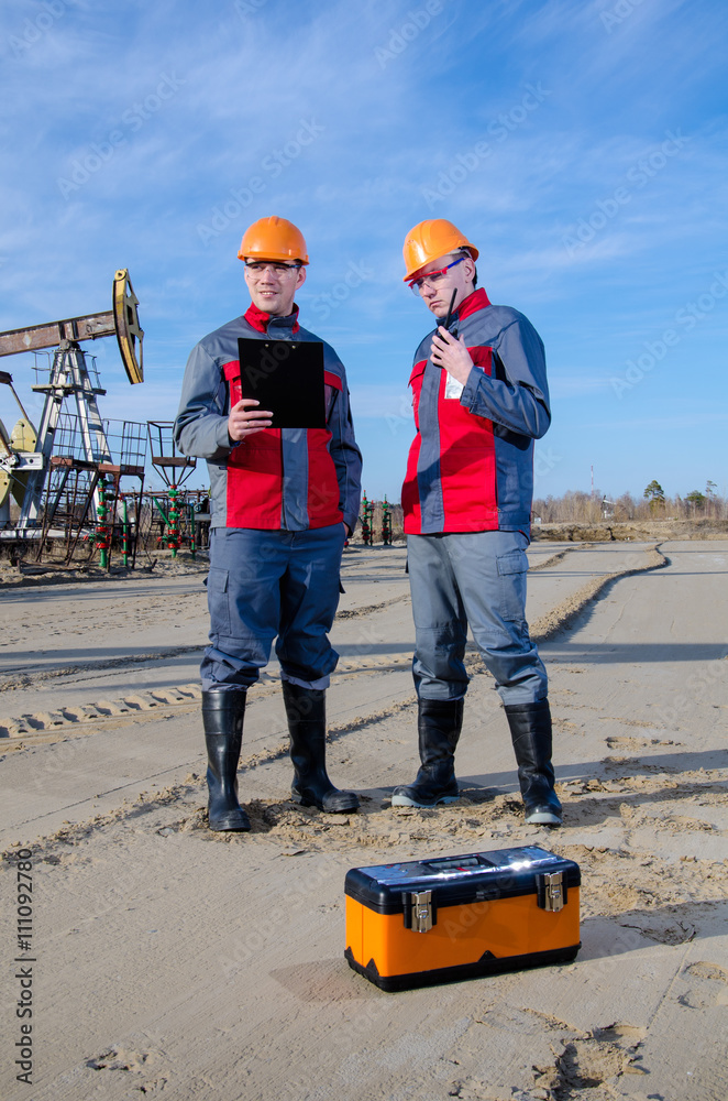 Workers in the oilfield, one holding radio in his hand. Pump jack ...