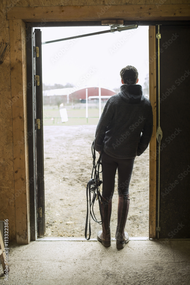 Full length rear view of young man with bridle leaning in doorway of ...