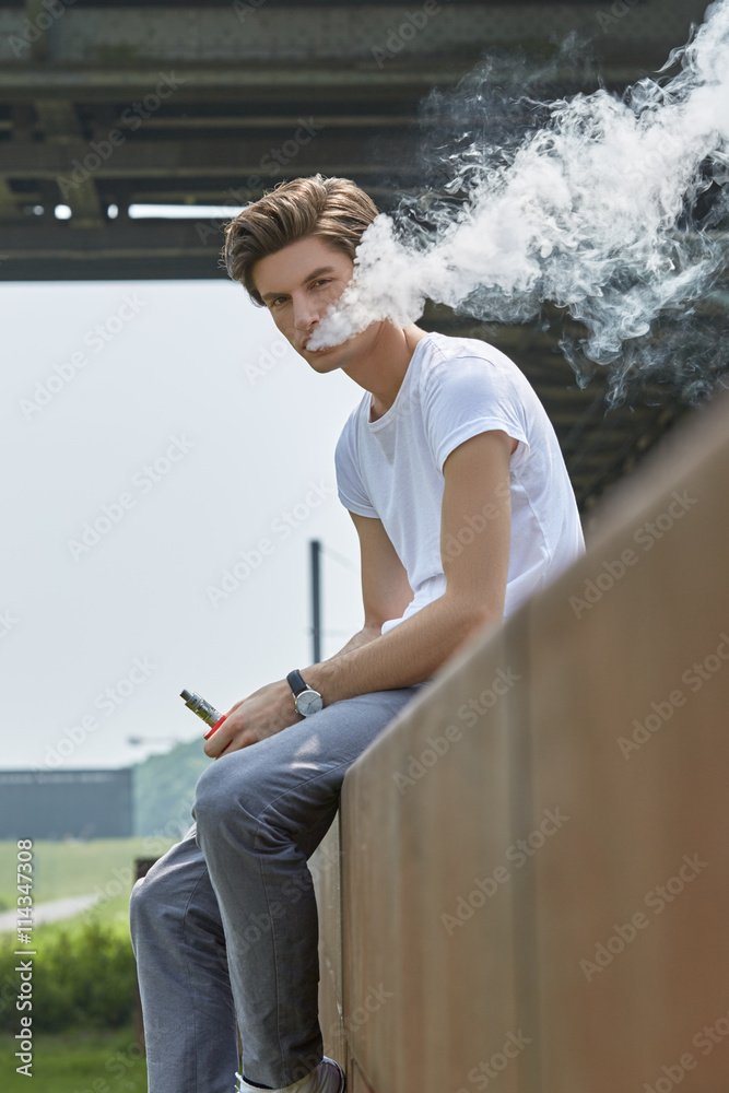 Foto de Junger Mann in T-Shirt und Jeans sitzt e-Zigarette rauchend auf ...