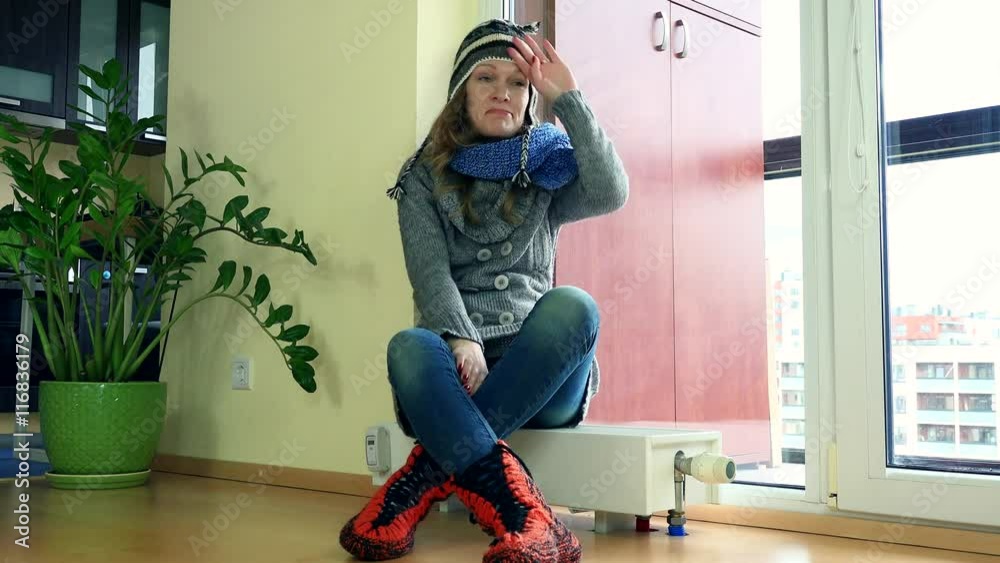 Cheerful girl strip warm clothes sitting on heater near balcony window ...