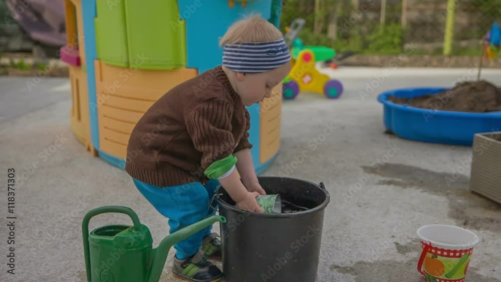 A toddler is scooping a cup of water from a bucket that is lying on the ...