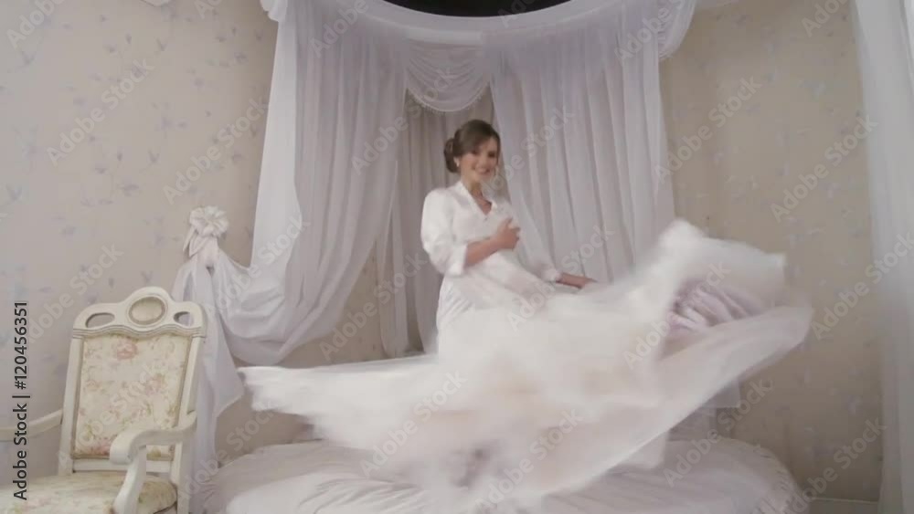 A happy bride with makeup and hairstyle in a lacy dressing gone jumps on the bed holding her ...