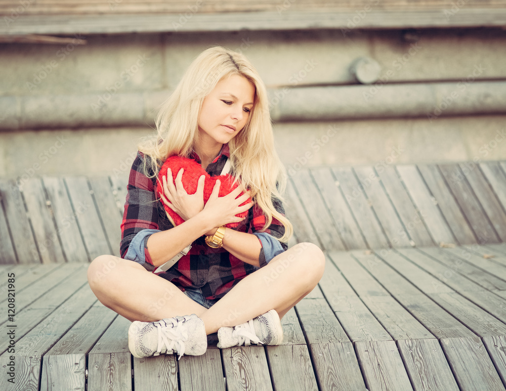 Sad lonely girl sitting on wood planks and hugging a big red heart ...
