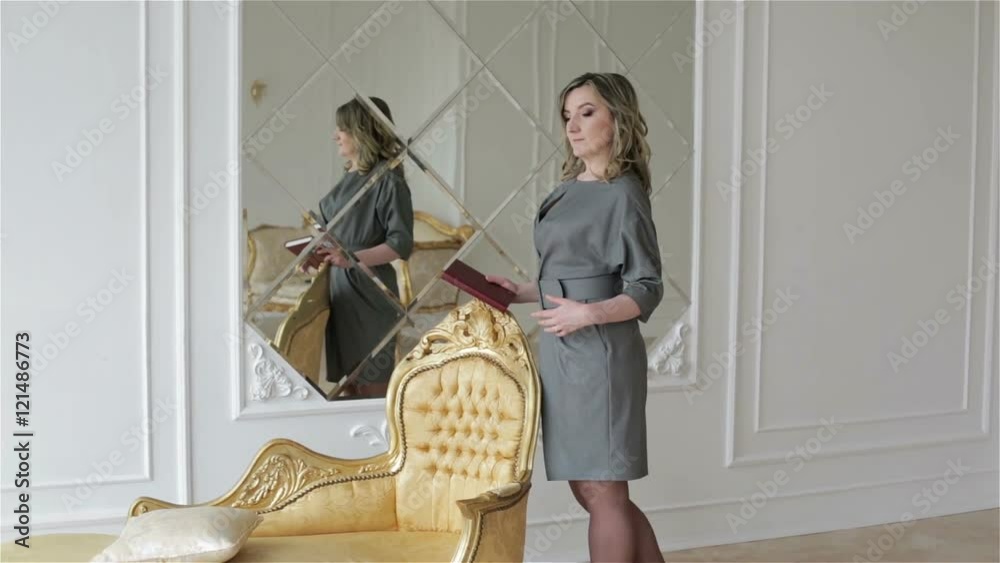 A young woman in grey dress is standing near the golden chair in studio ...