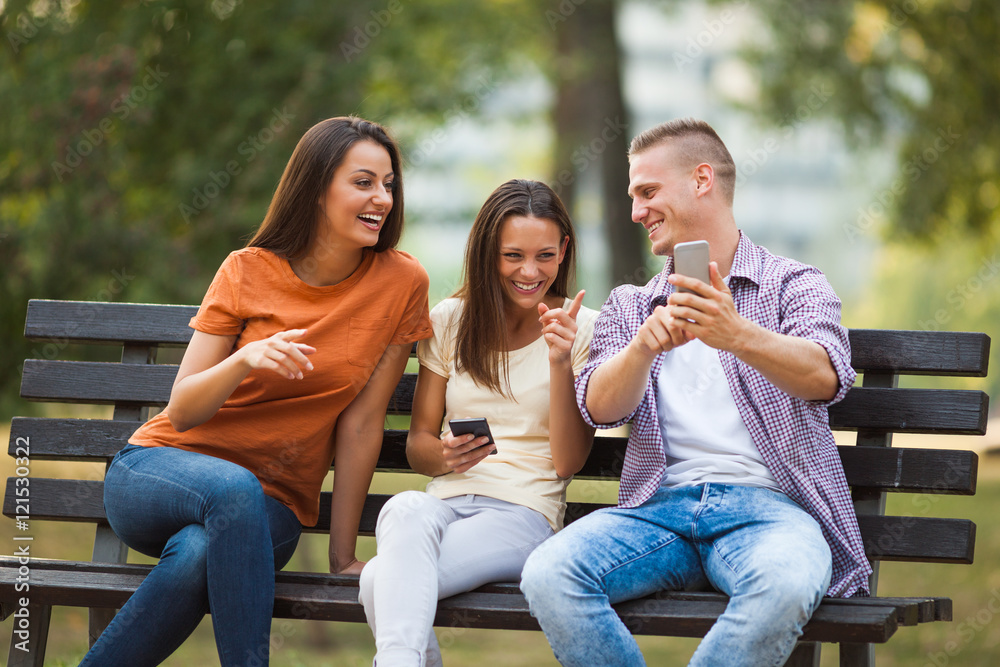 Three friends are sitting on bench in park and using smartphones. Stock ...