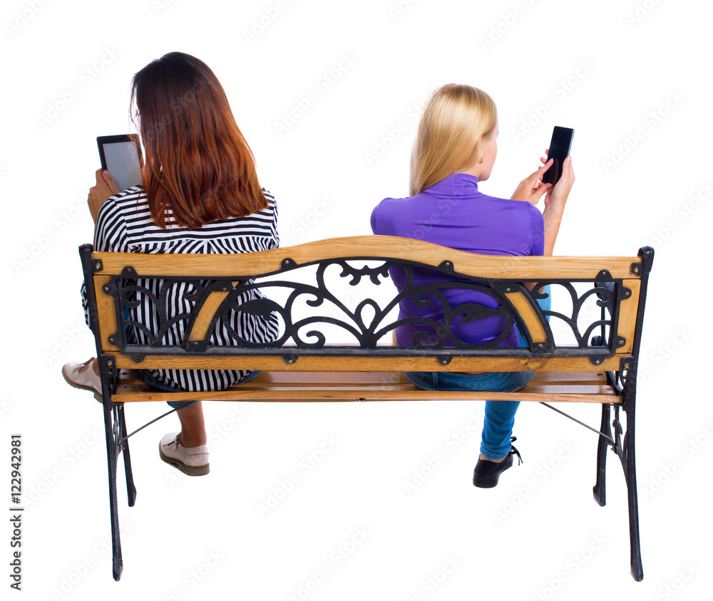 back view of two women sitting on bench and looks at the screen of the ...