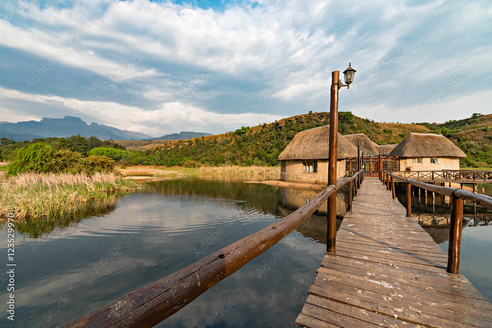 Wooden bridge over dam, leading to thatched building, alongside a park ...