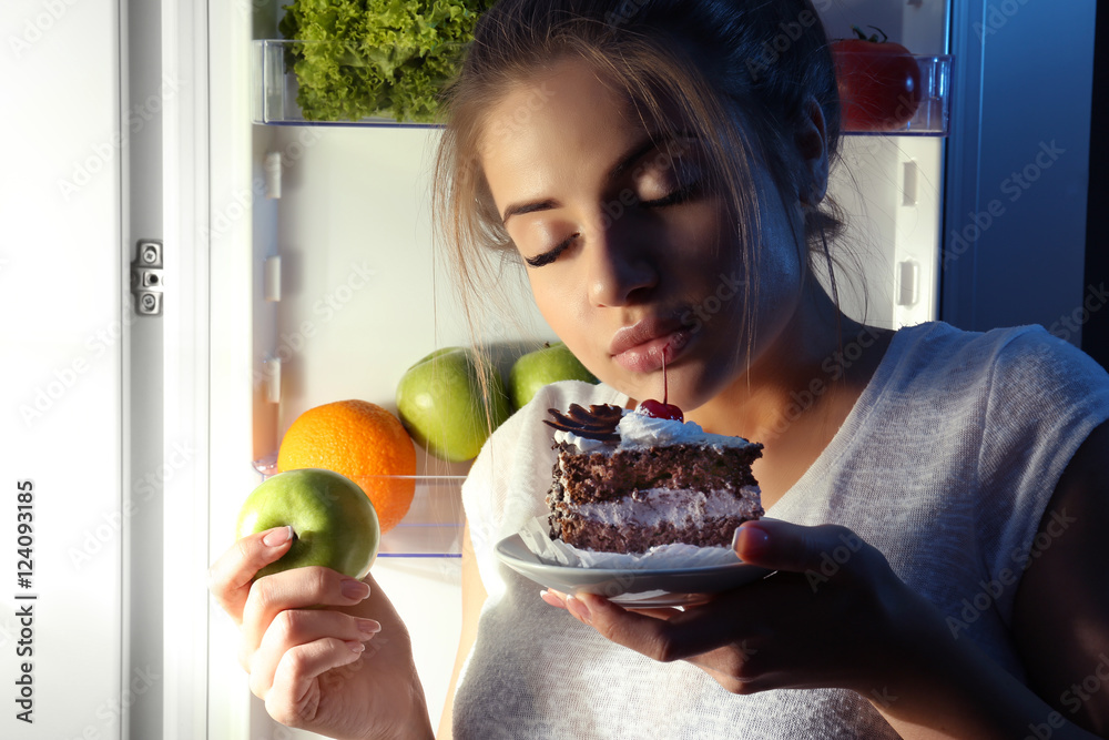 Beautiful girl eating cake and apple at night. Unhealthy food concept ...