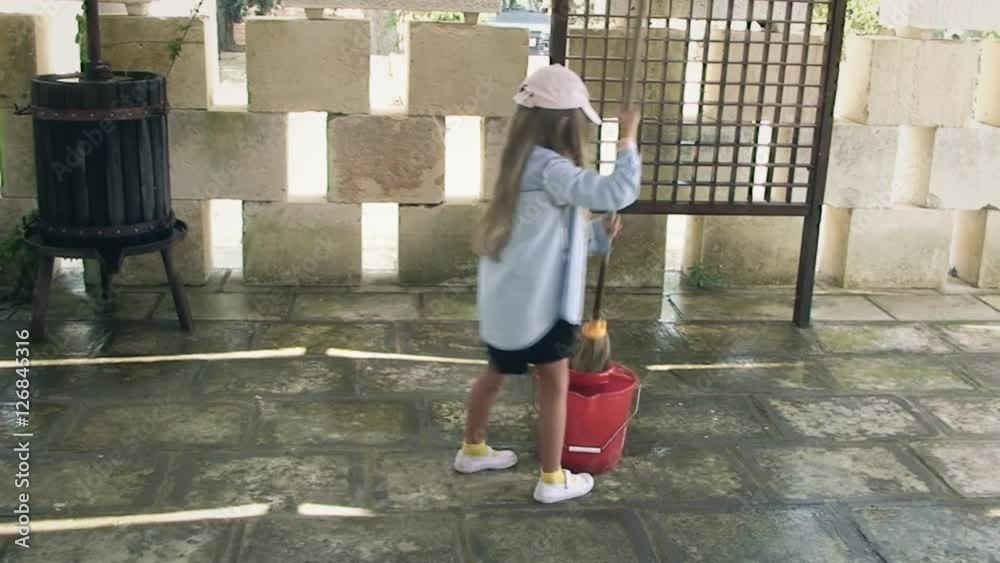 A little girl washing and cleaning up a stone floor with a mop. Outside ...
